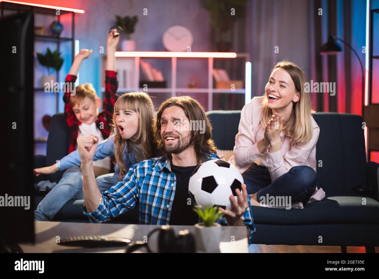 Positive young parents with little son and daughter sitting on couch and watching soccer games ...