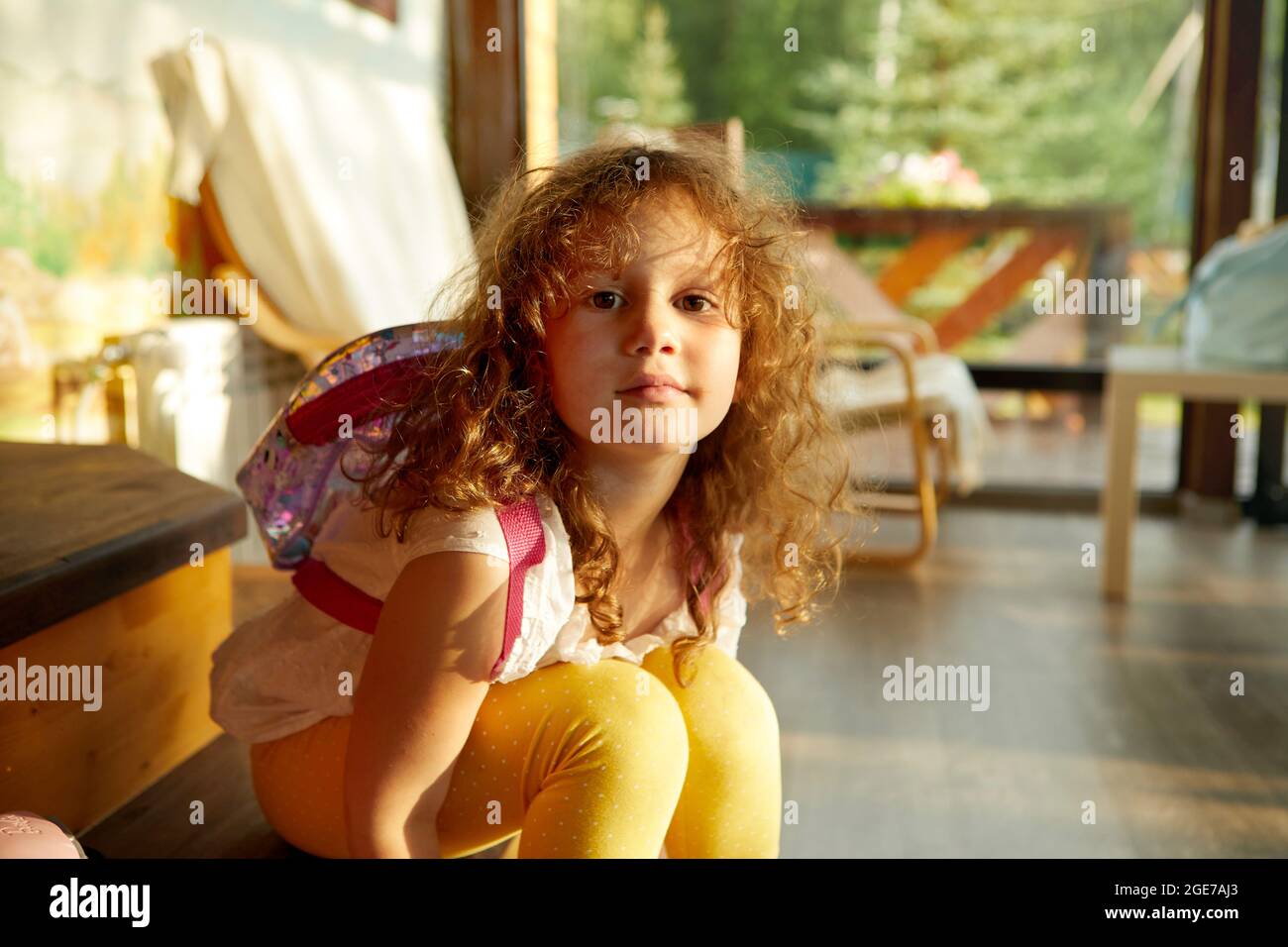 Adorable girl with backpack bending forward and looking at camera while ...