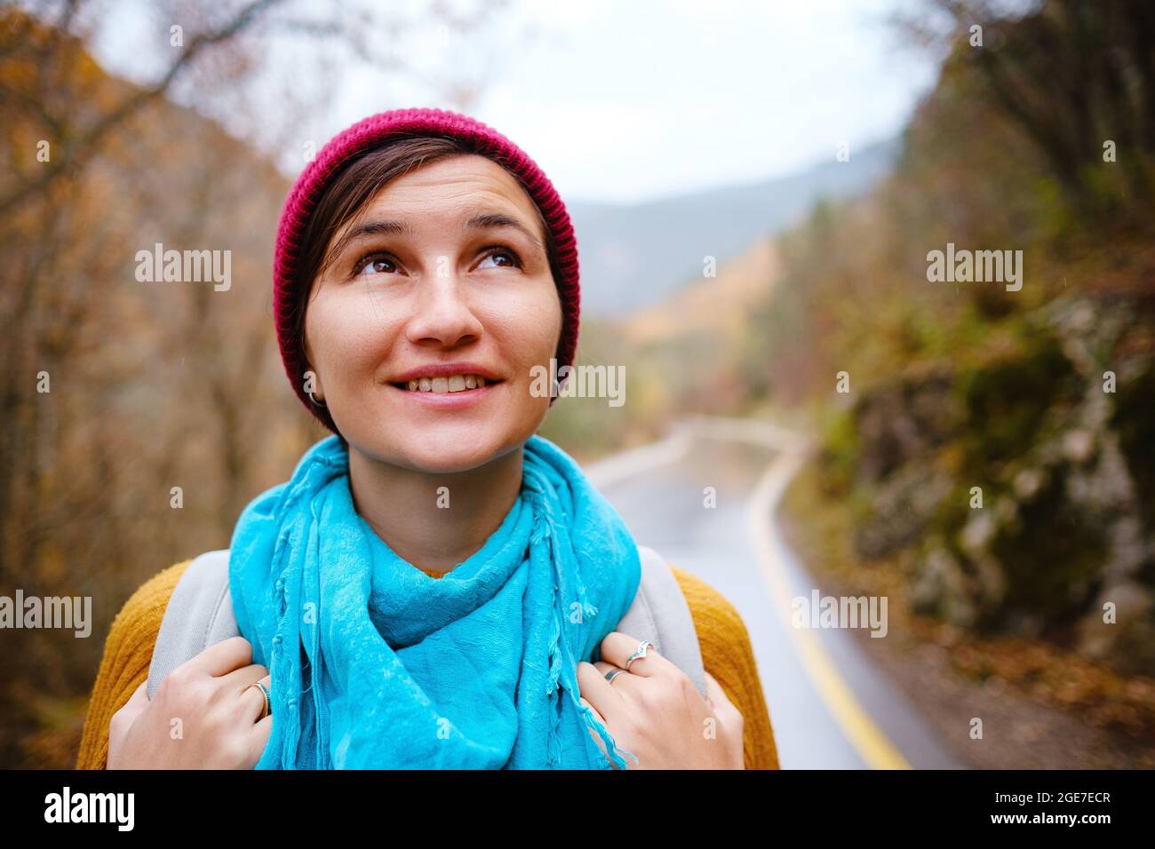 beautiful female hipster in a red hat, yellow sweater and blue scarf walks in the autumn nature ...