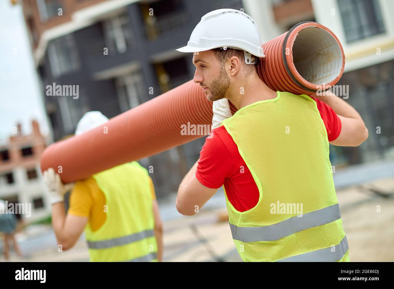 Two workers in protective helmets carrying a corrugated drain pipe ...