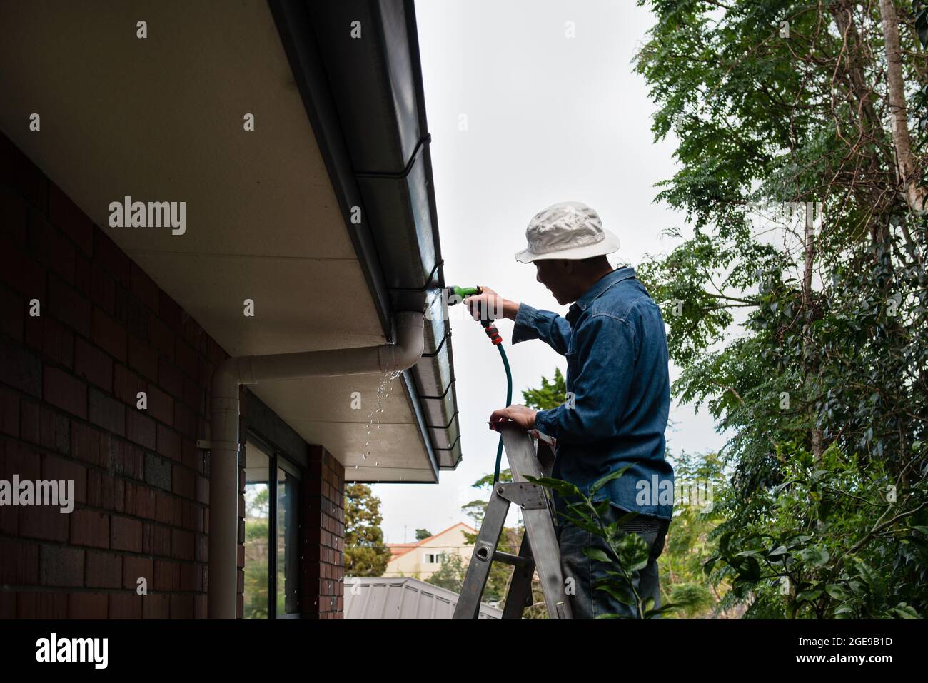 Man standing on the ladder and washing the gutter using a garden hose ...