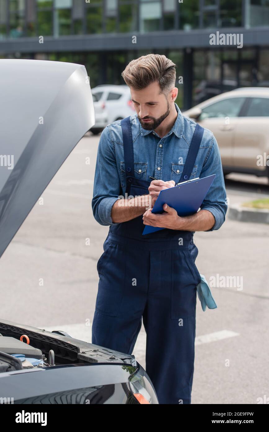 Mechanic writing on clipboard while looking at engine of car outdoors ...