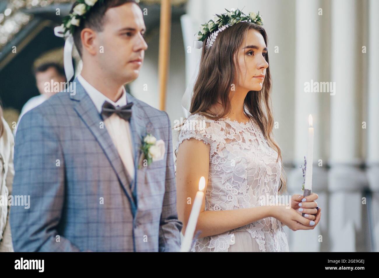 Stylish bride and groom in floral crowns holding candles during holy ...