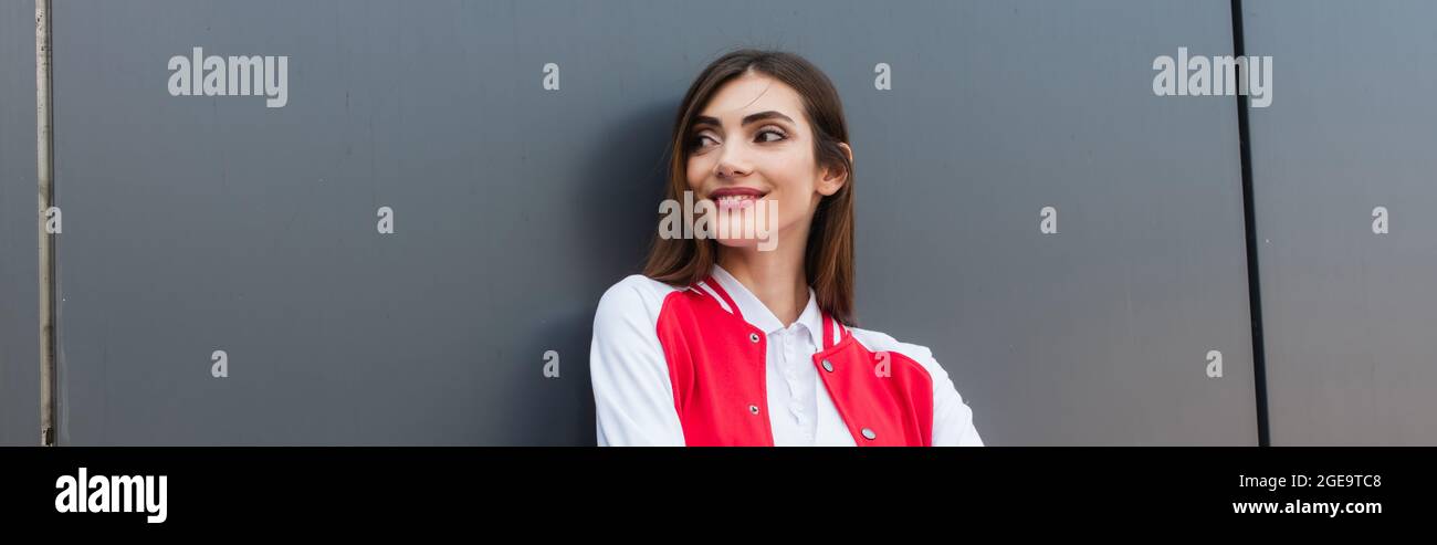 happy woman in black and white jacket looking aside near grey wall ...