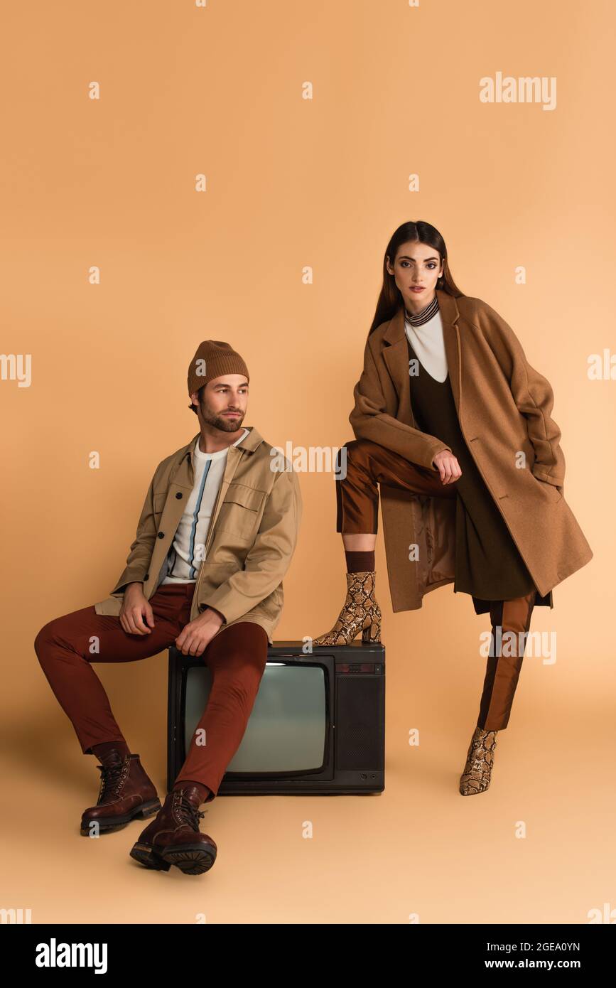 woman in stylish autumn coat looking at camera near trendy man sitting ...