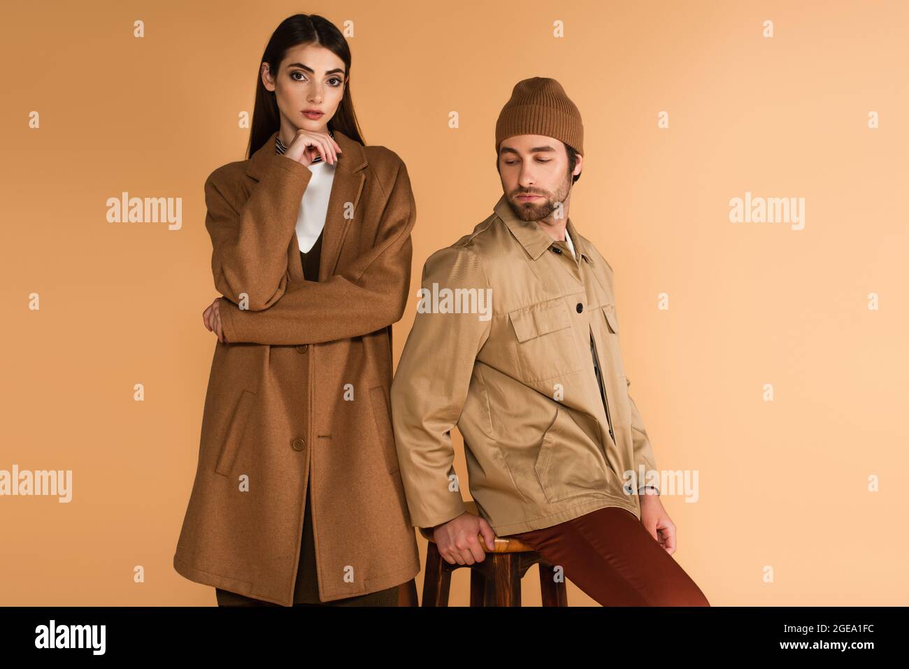 woman in brown coat standing near stylish man sitting on stool isolated ...