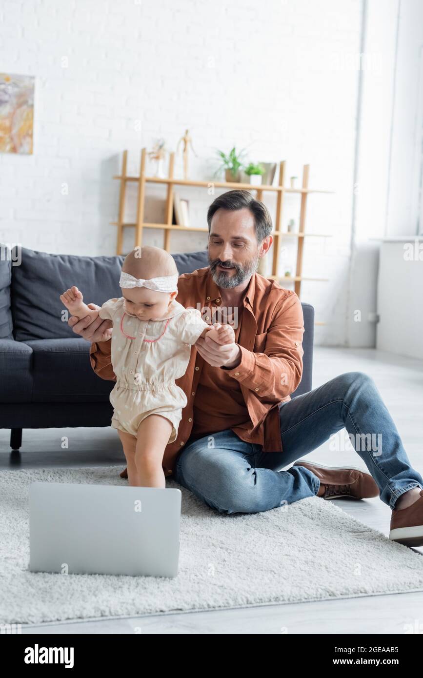 Smiling man holding hands of toddler daughter near laptop on carpet ...