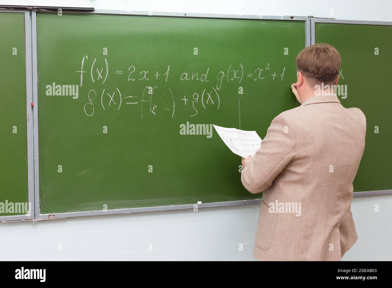 Mathematics teacher writes in chalk on a blackboard with his back to ...