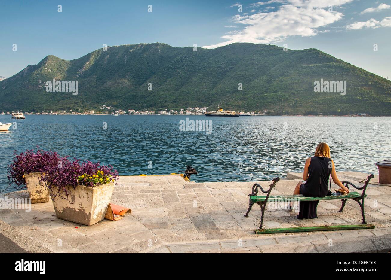 A young woman sits on a waterside bench overlooking Kotor Bay at the ...