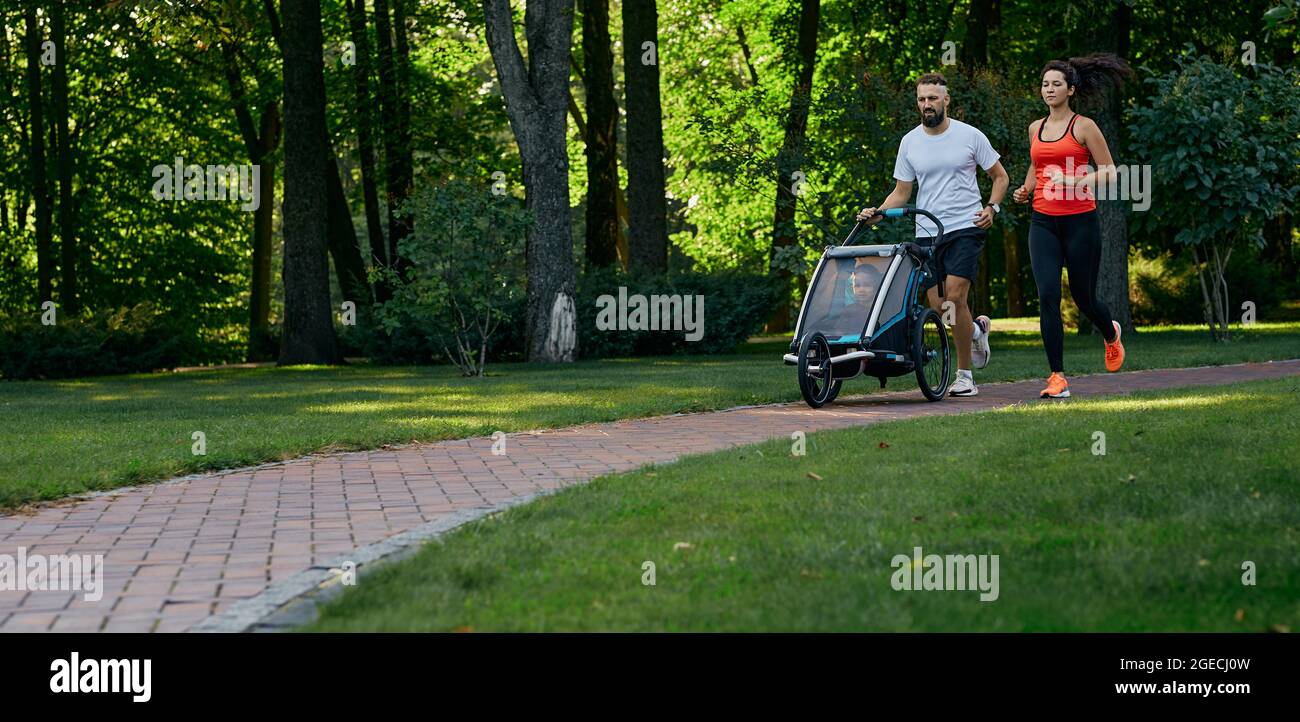 Running couple with child in jogging stroller in public park at sunrise ...