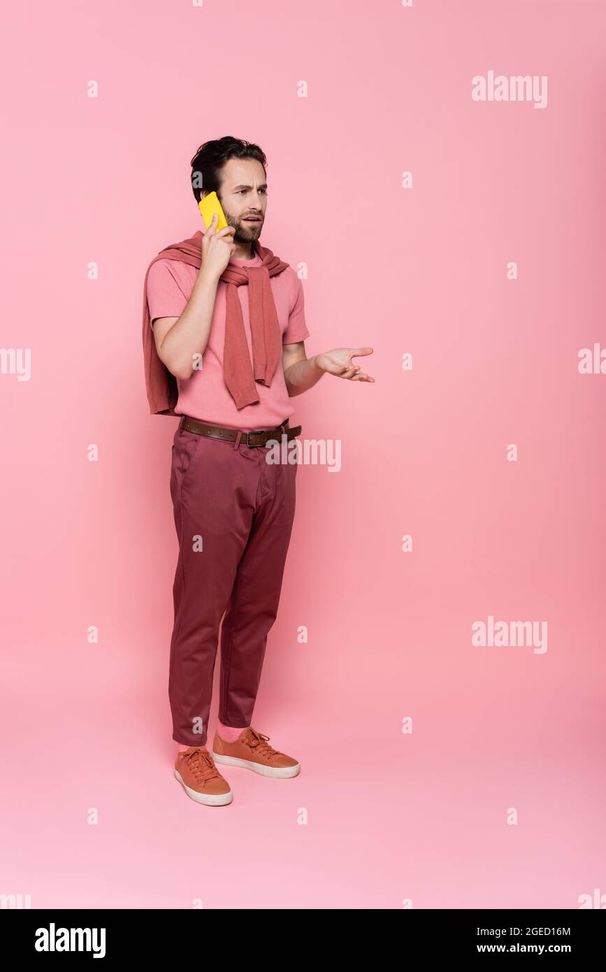 Full length of confused man talking on mobile phone on pink background ...