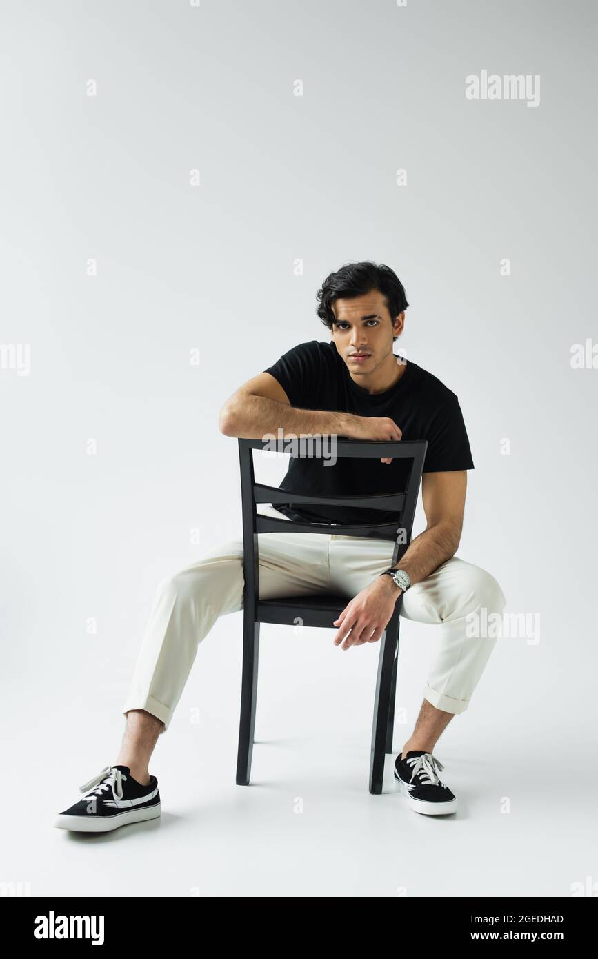 full length of young man looking at camera and sitting on chair on grey ...