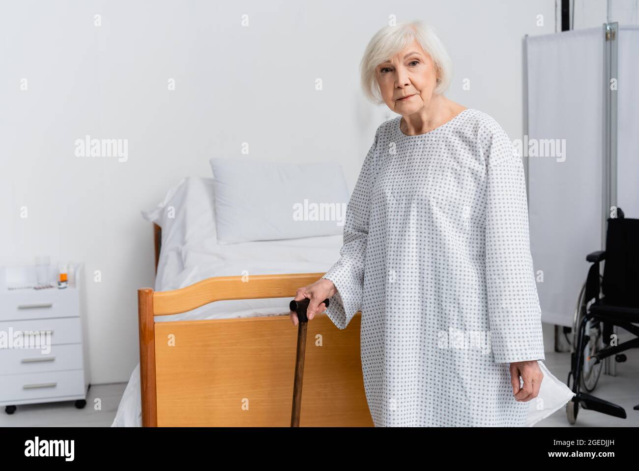 Elderly patient with walking cane looking at camera near bed and ...