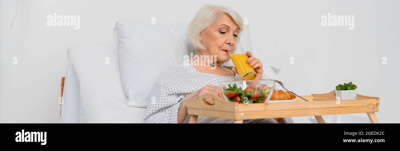 Senior patient drinking orange juice near croissant and salad on tray ...