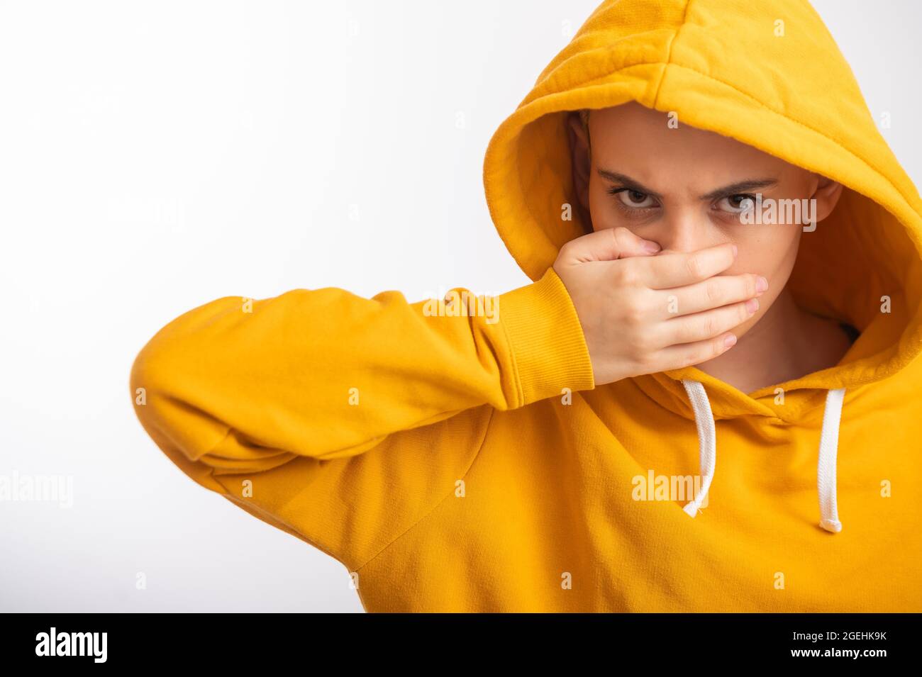 Young woman in ocher hood covers her face with hand on white background ...