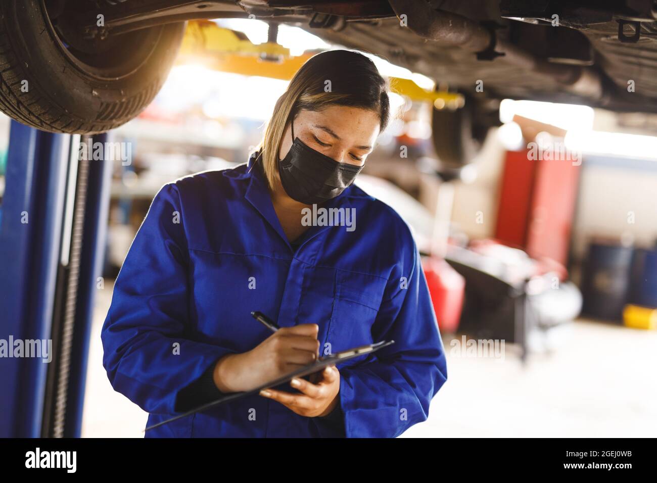 Mixed race female car mechanic wearing face mask and overalls, making ...