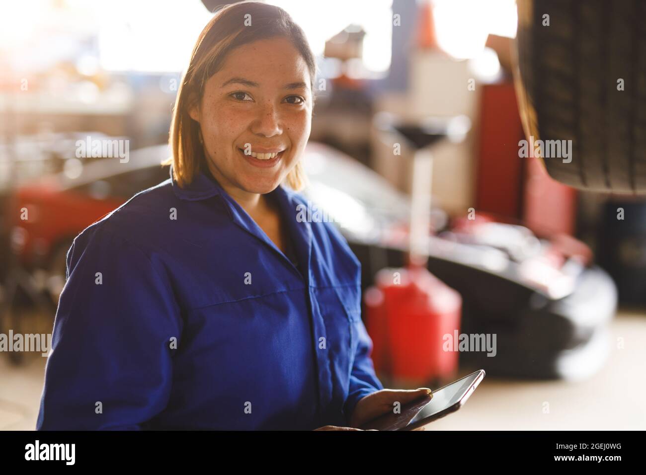 Smiling mixed race female car mechanic wearing overalls, holding tablet ...