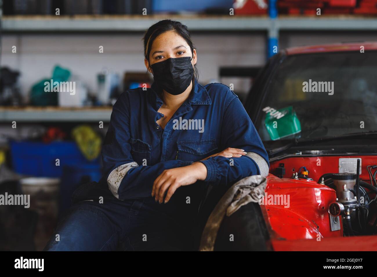 Mixed race female car mechanic wearing face mask and overalls, looking ...