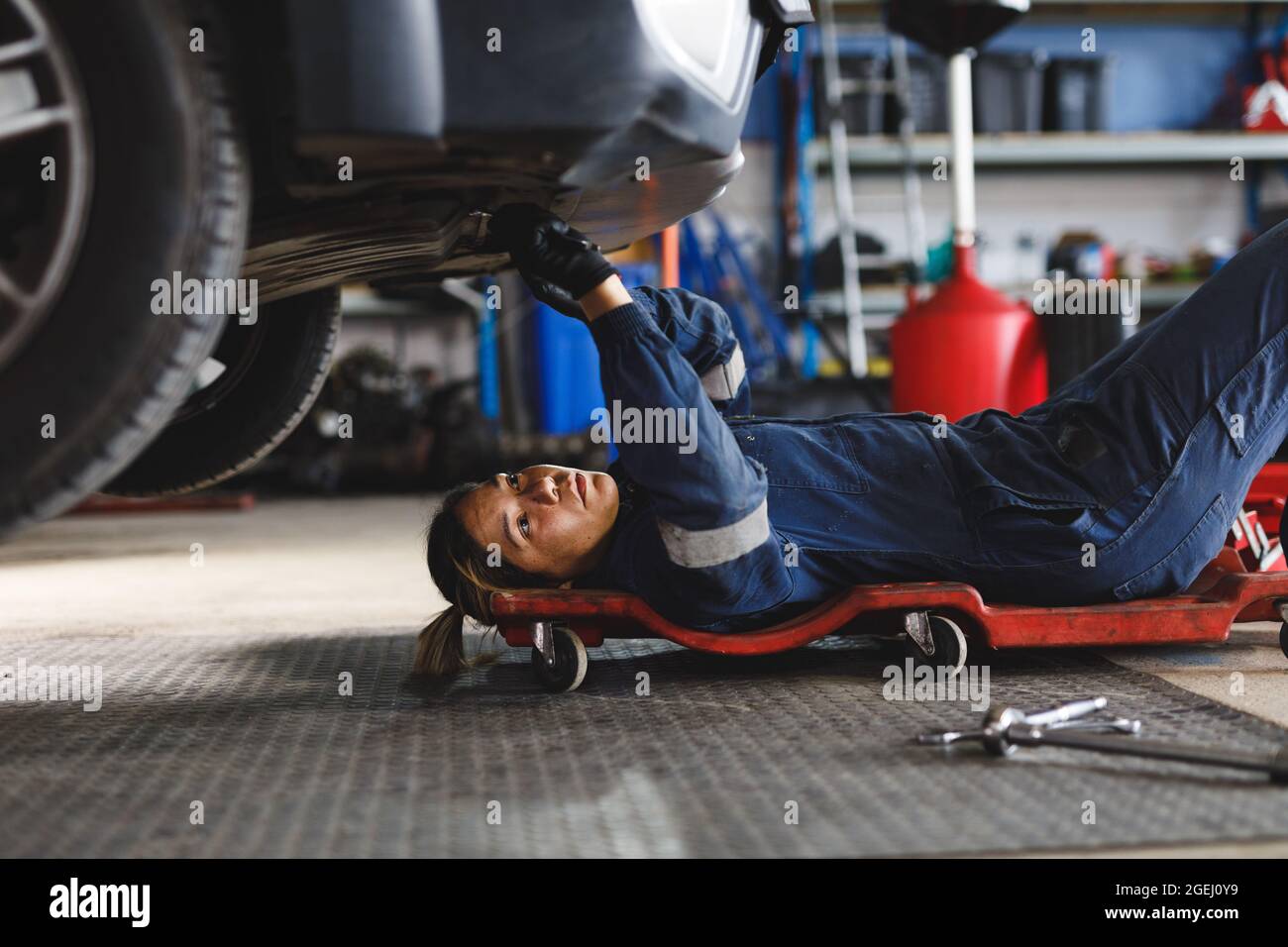 Mixed race female car mechanic wearing overalls, lying on a board ...