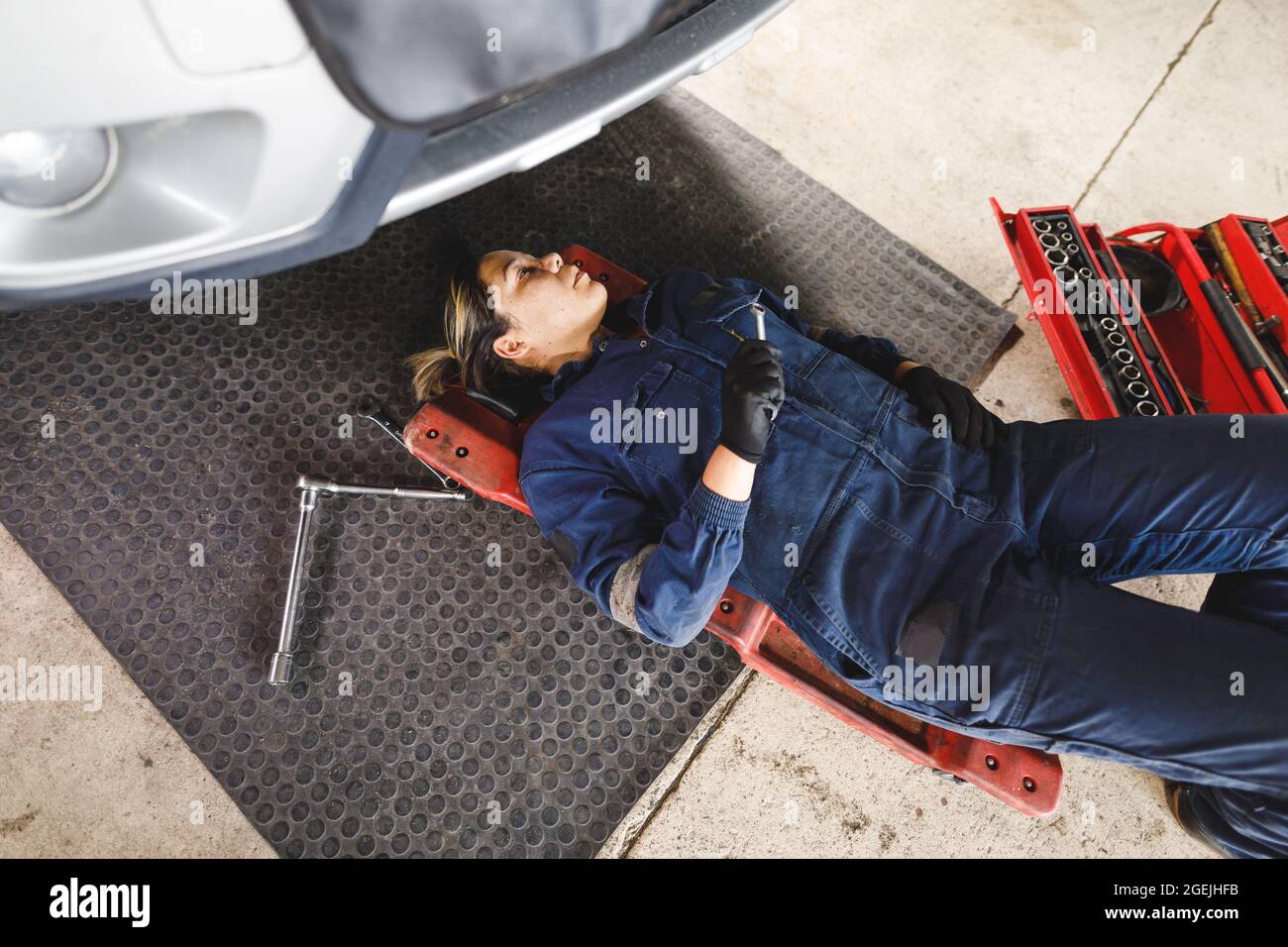 Mixed race female car mechanic wearing overalls, lying on a board ...