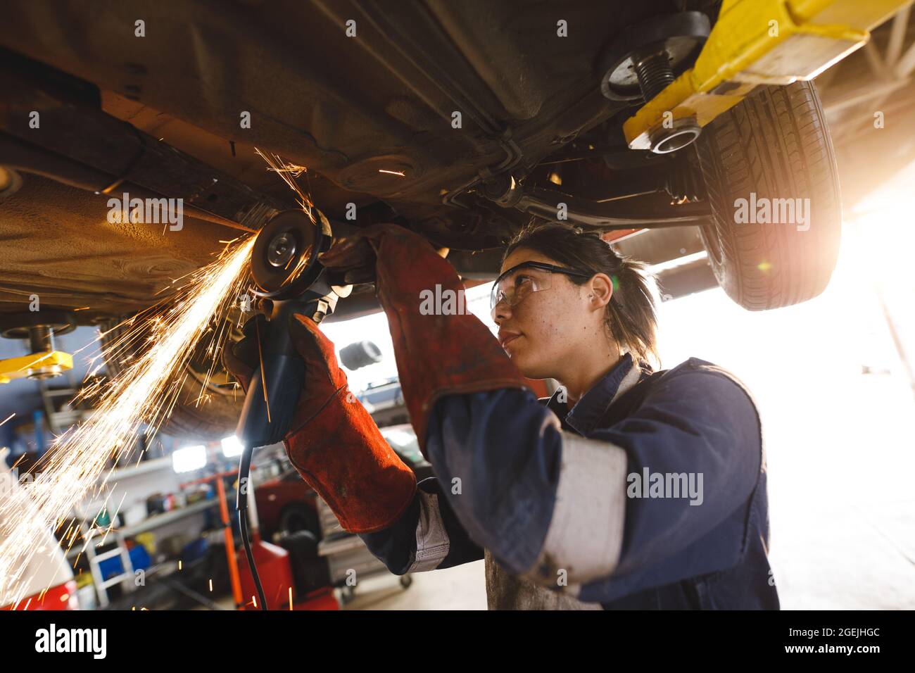 Mixed race female car mechanic wearing overalls, using angle grinder ...