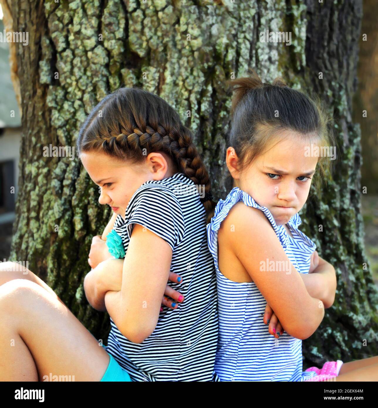 Two little girls sit back-to-back, outside besides a tree. They both ...