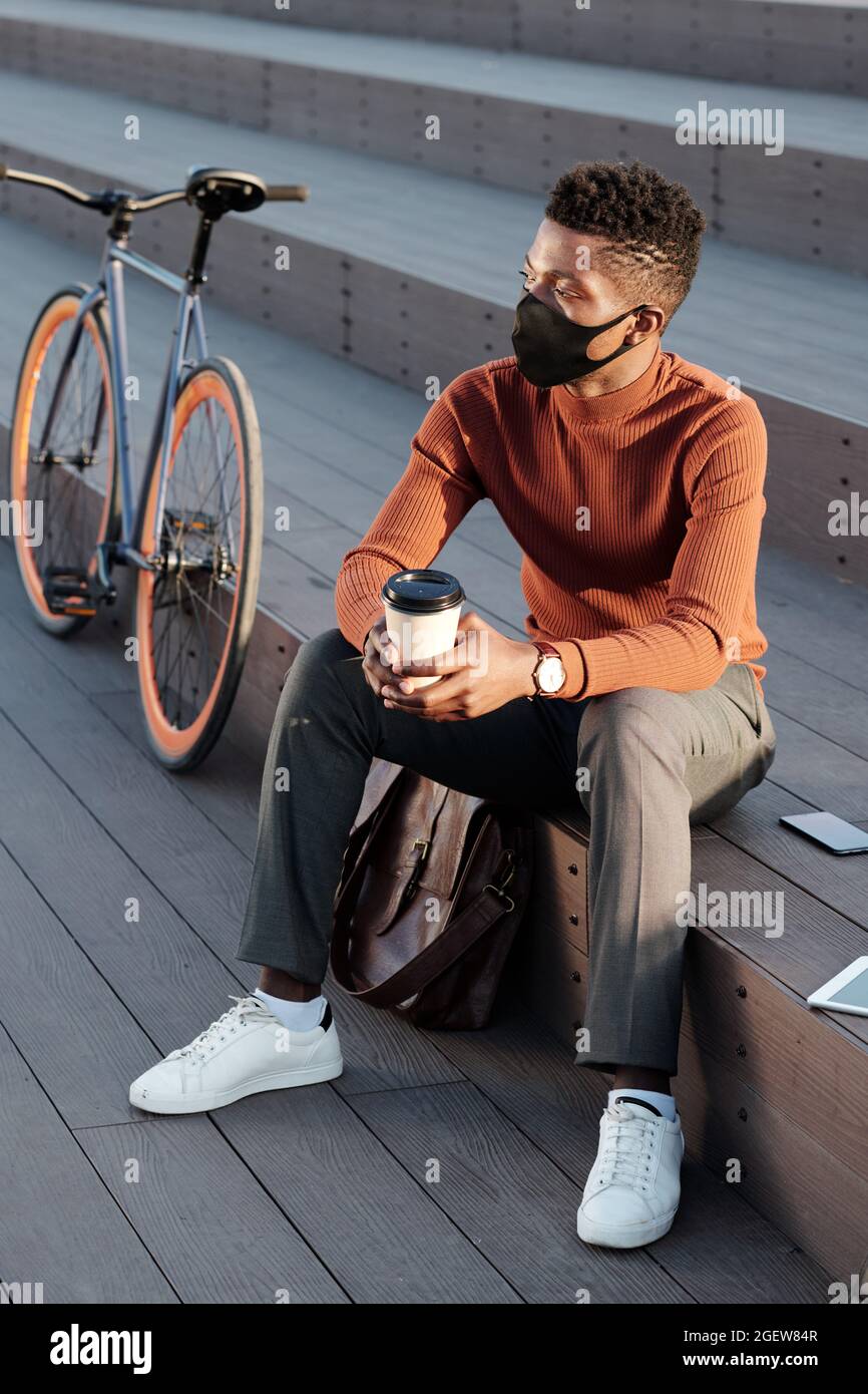 Young restful businessman with drink sitting on staircase while having ...