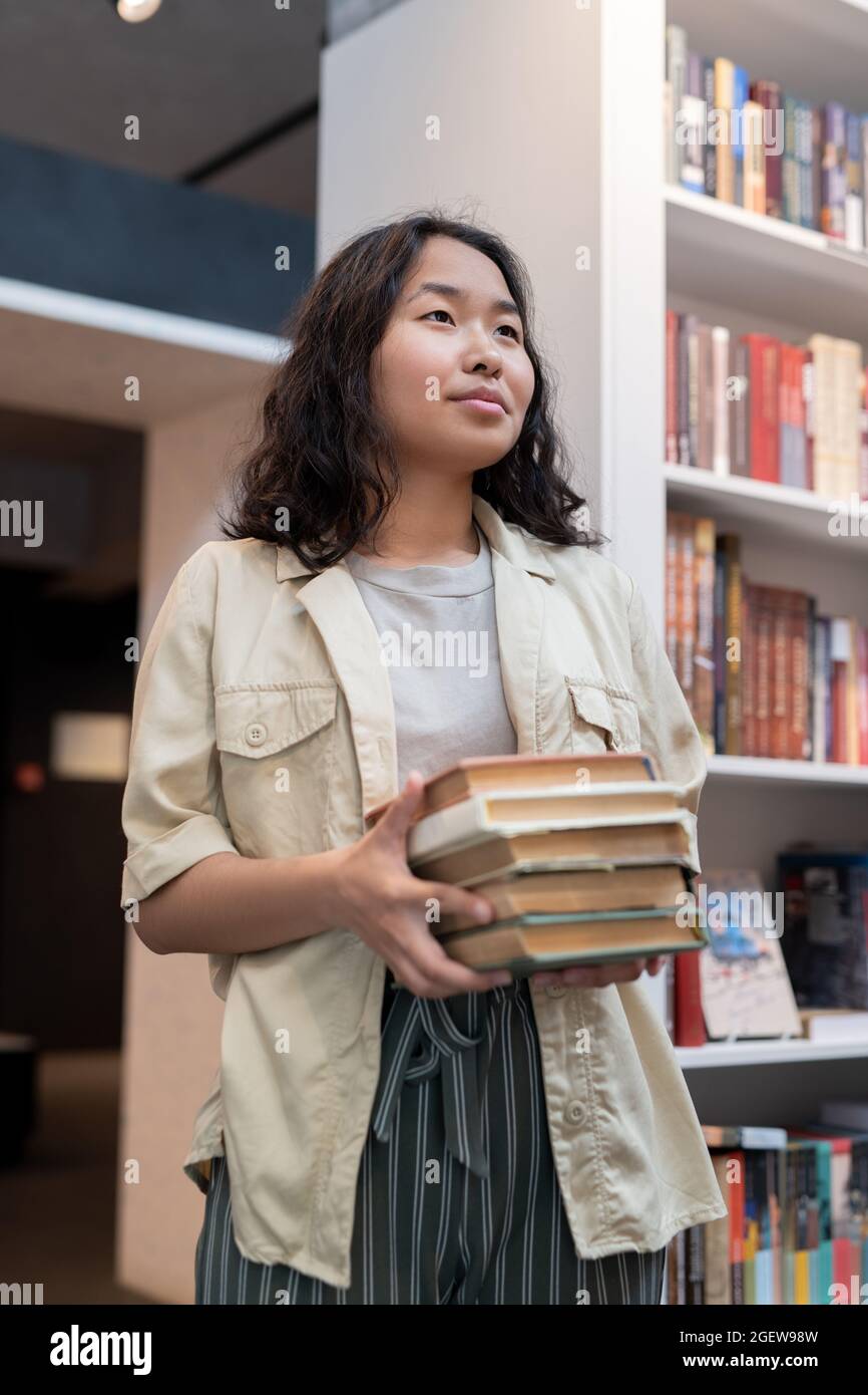 Contemporary student with stack of books standing against shelf in ...