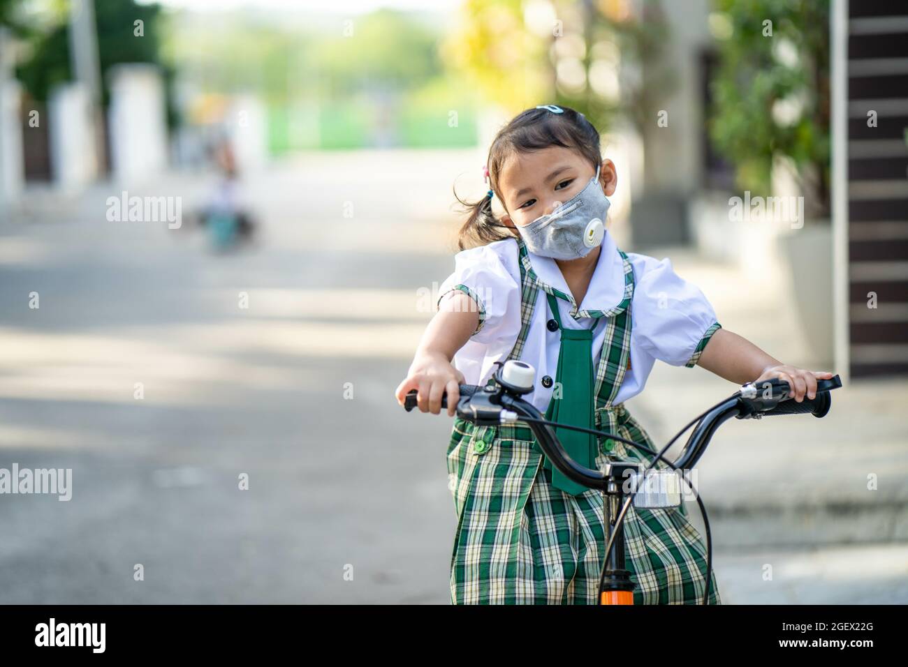 Cute little Thai girl in a school uniform and a face mask riding a bike ...
