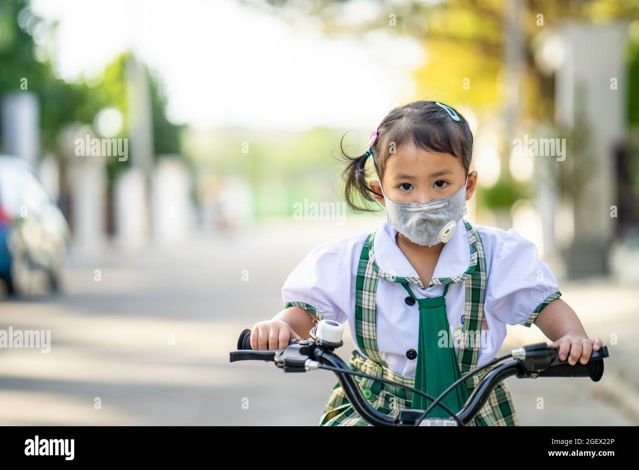 Cute little Thai girl in a school uniform and a face mask riding a bike ...
