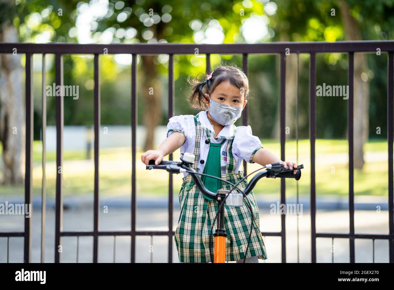 Cute little Thai girl in a school uniform and a face mask riding a bike ...