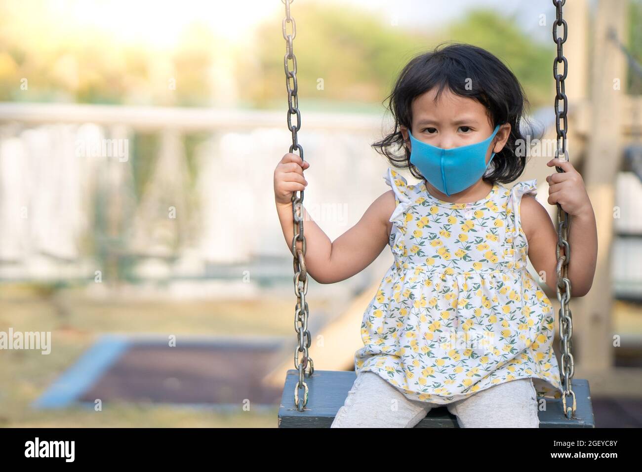 A little girl wearing a medical disposable face mask sitting at chain ...