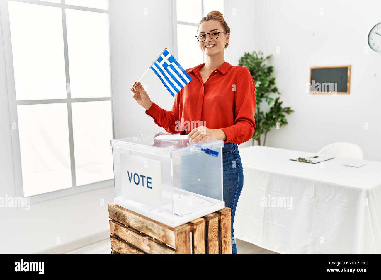 Young brunette woman putting envelop in ballot box holding greece flag ...