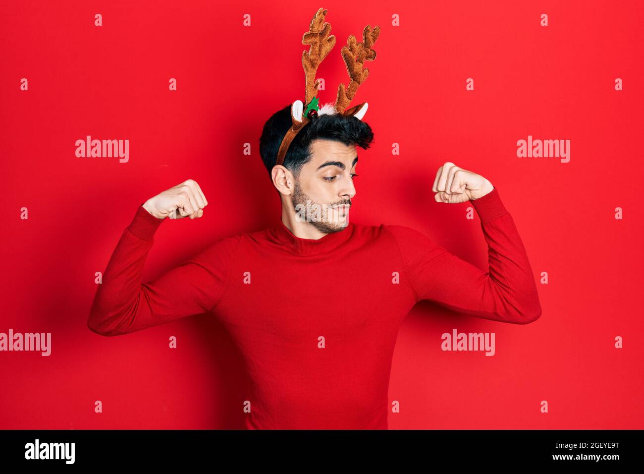 Young hispanic man wearing cute christmas reindeer horns showing arms ...