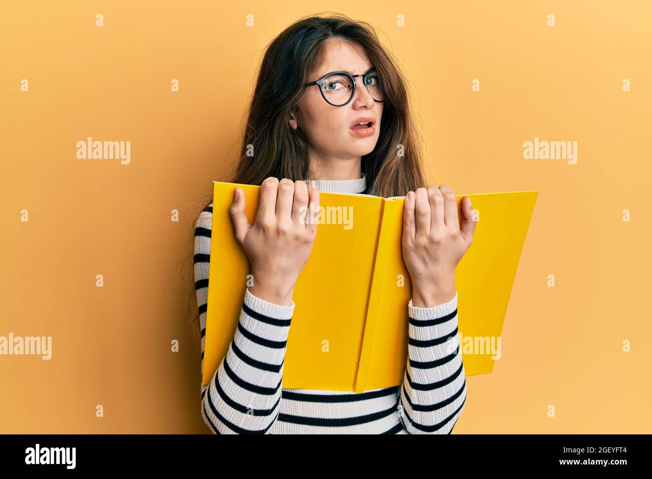 Young caucasian woman reading a book wearing glasses in shock face ...