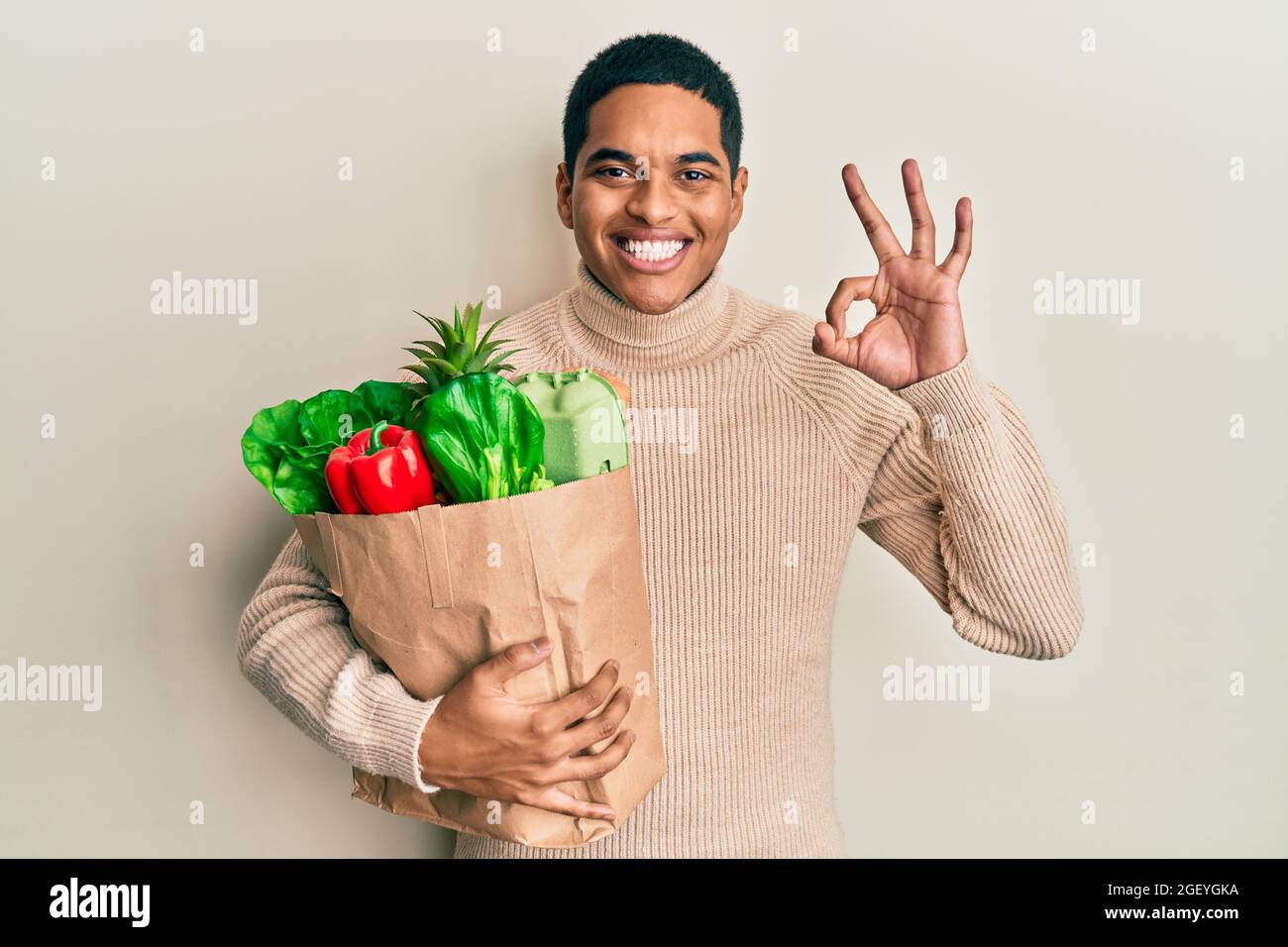 Young handsome hispanic man holding paper bag with groceries doing ok ...
