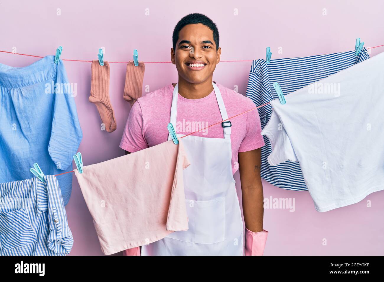 Young handsome hispanic man wearing cleaner apron holding clothes on ...