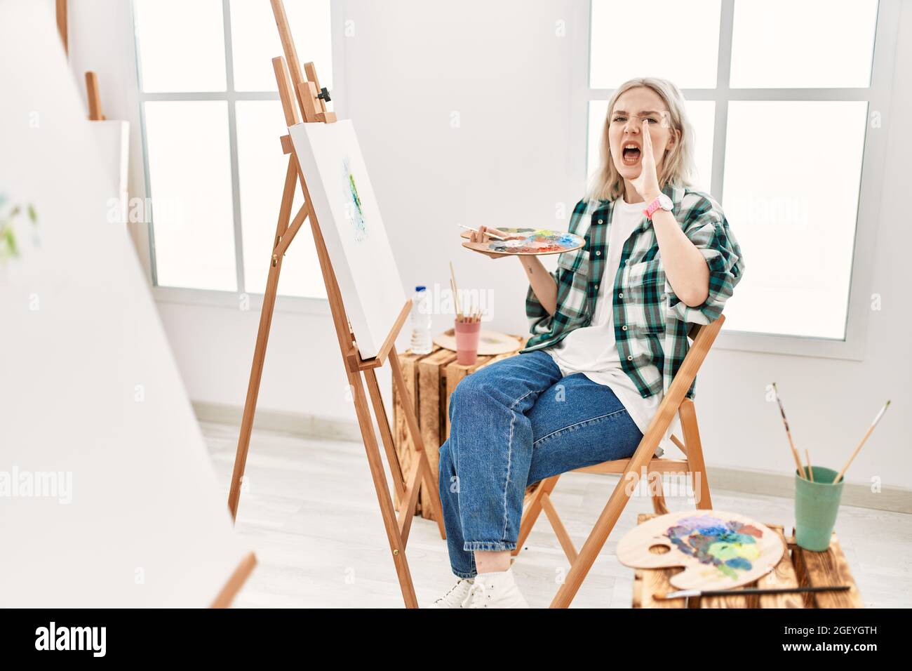 Young artist woman painting on canvas at art studio shouting and ...
