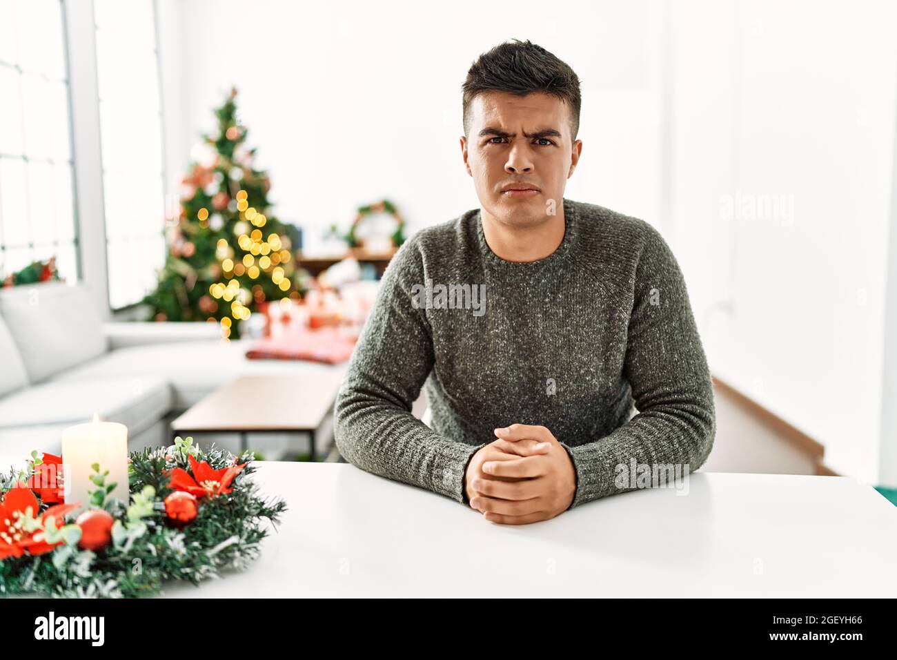 Young hispanic man sitting on the table by christmas tree skeptic and ...
