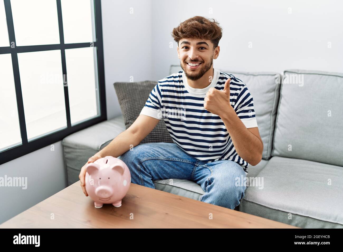 Young arab man holding piggy bank smiling happy and positive, thumb up ...