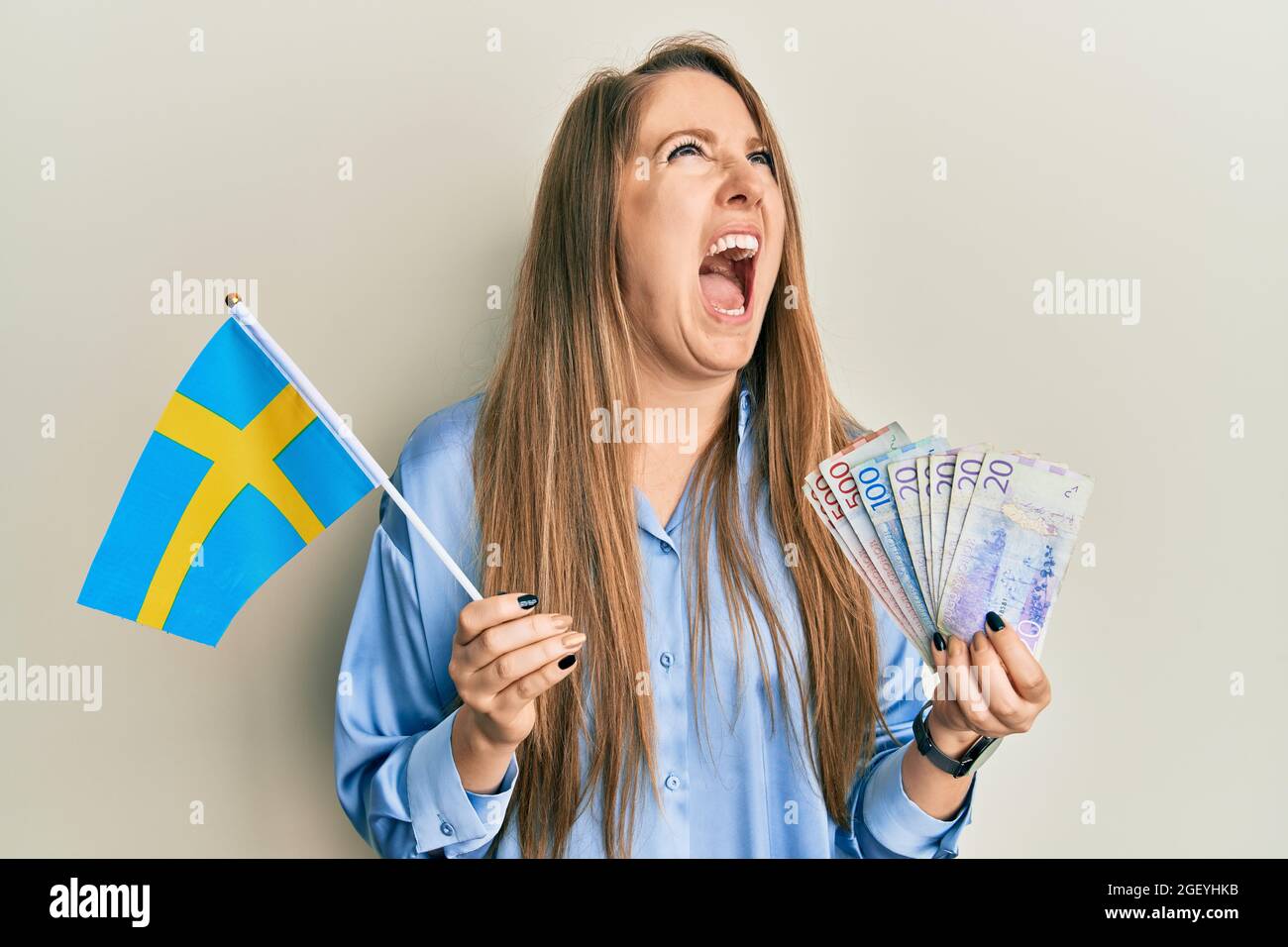 Young blonde woman holding sweden flag and krone banknotes angry and ...