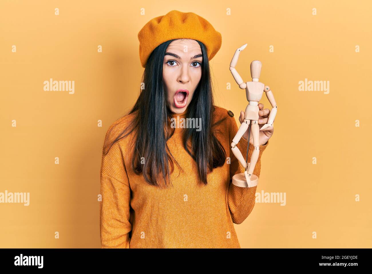 Young brunette woman holding small wooden manikin wearing beret scared ...