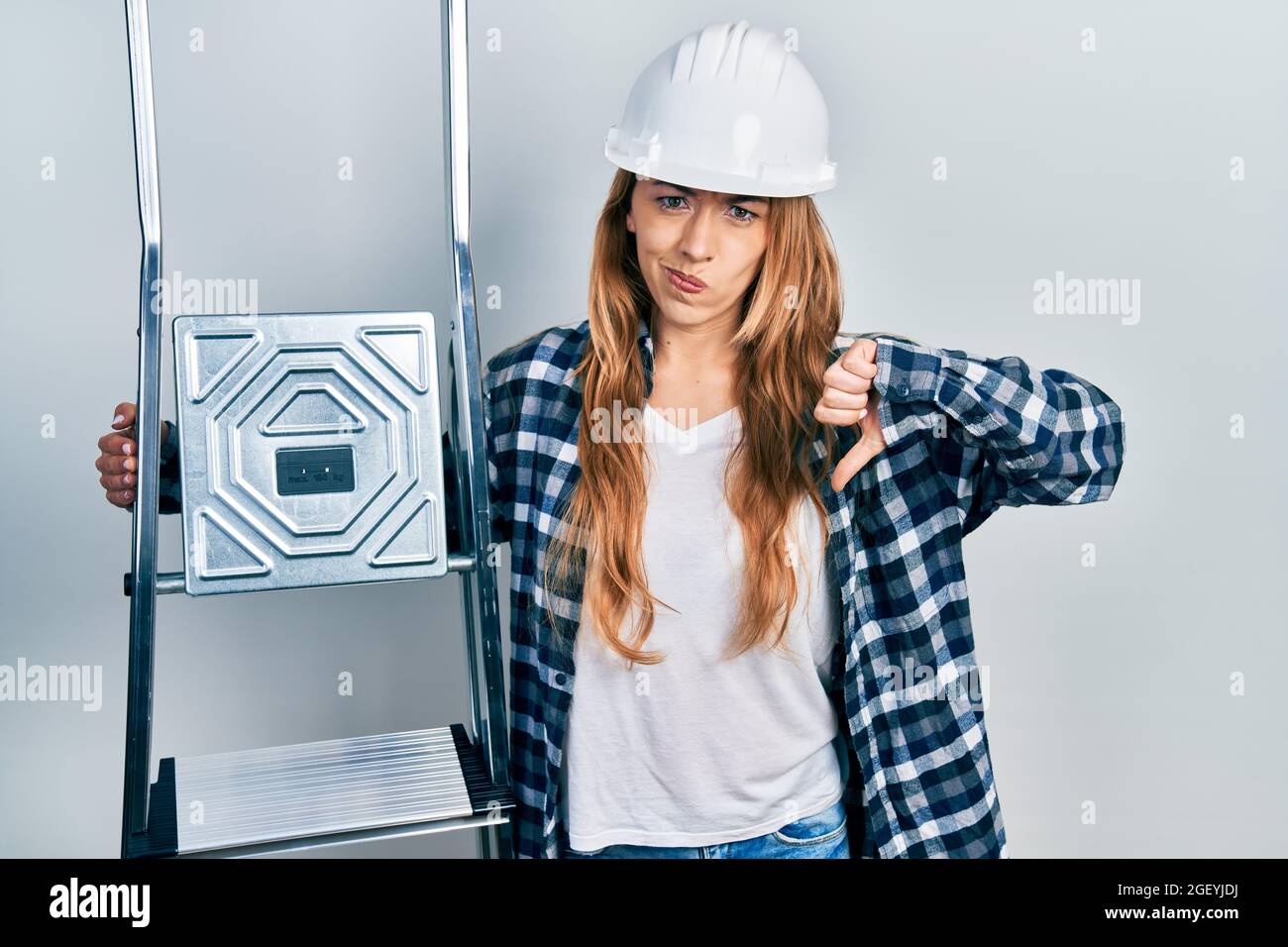 Young caucasian woman wearing hardhat holding ladder with angry face ...