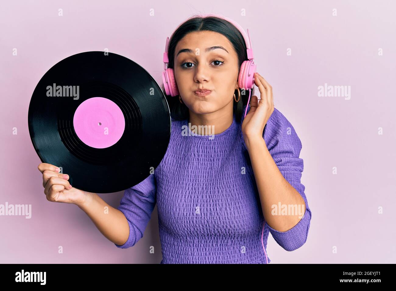 Young hispanic woman using headphones holding vinyl disc puffing cheeks ...