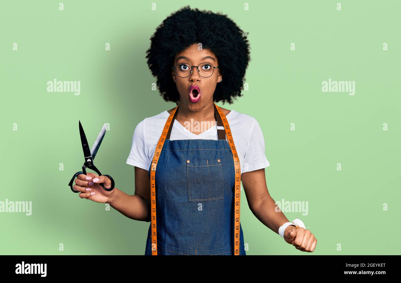 Young african american woman dressmaker designer wearing atelier apron holding scissors scared ...