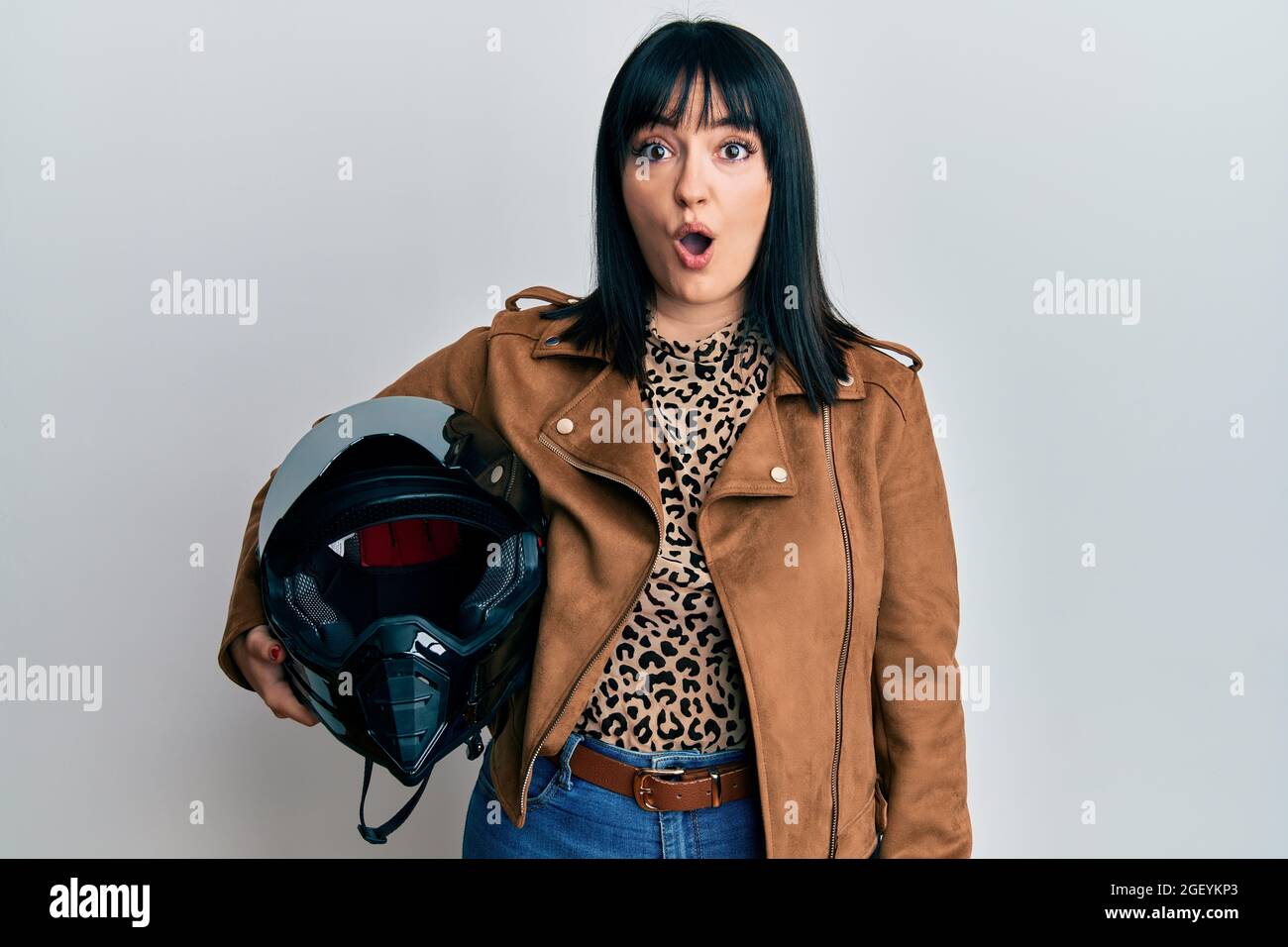 Young hispanic woman holding motorcycle helmet scared and amazed with ...