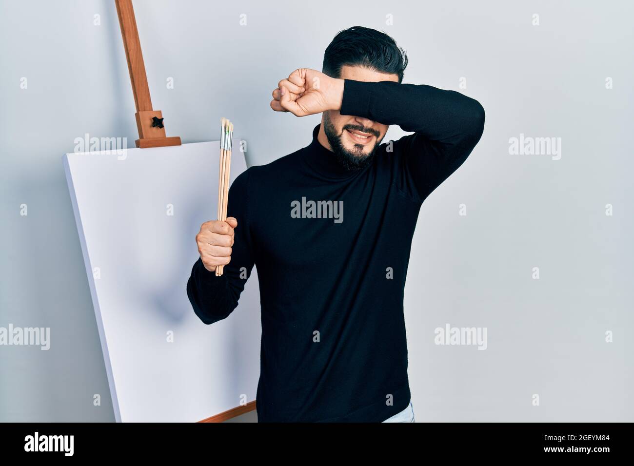 Handsome man with beard holding brushes close to easel stand smiling ...