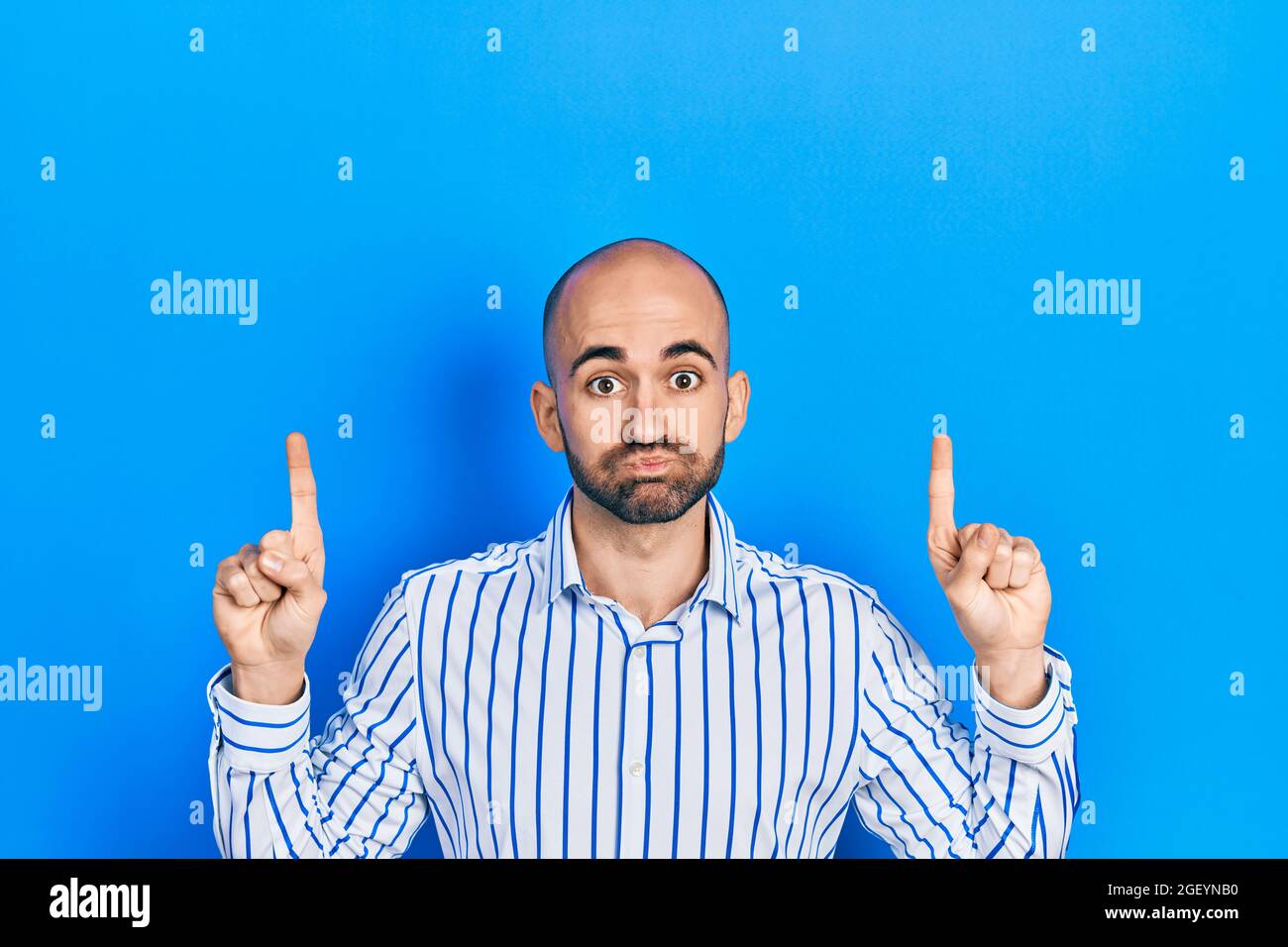 Young bald man pointing up with fingers puffing cheeks with funny face ...