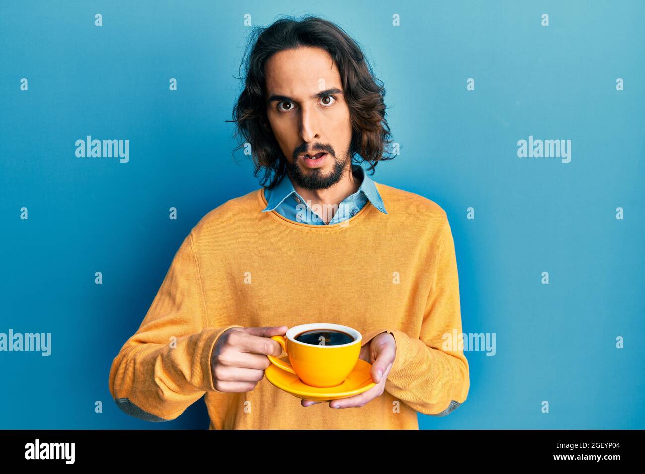 Young hispanic man drinking a cup of coffee in shock face, looking ...