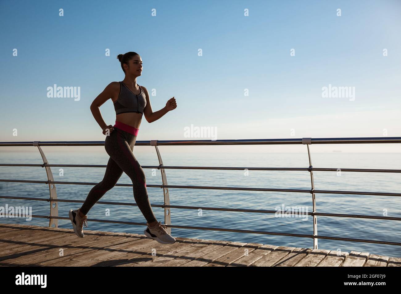 Side view of young fitness woman running along pier. Female athlete workout and jogging on the ...